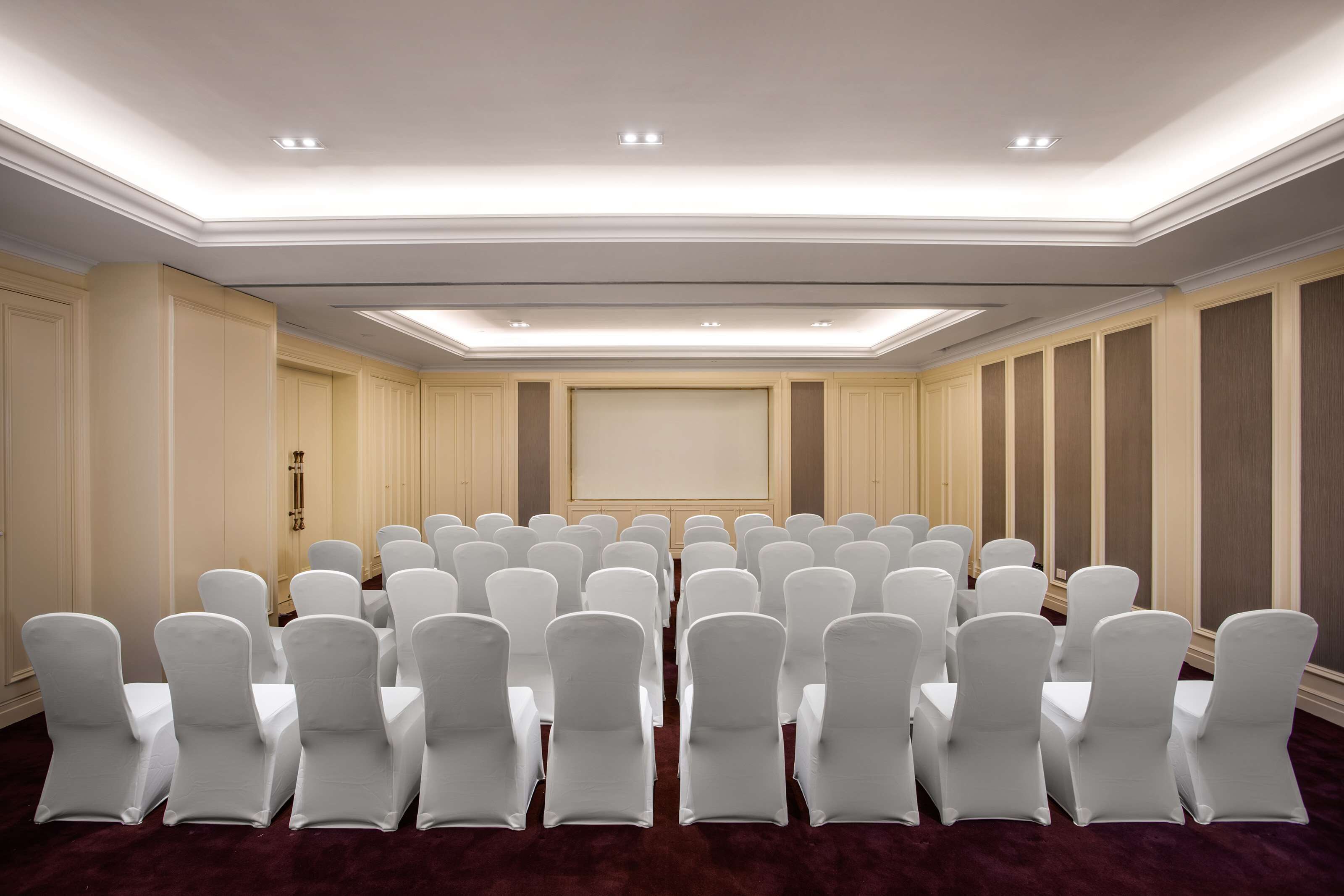 Uma sala de conferências com fileiras de cadeiras brancas, uma tela grande e iluminação embutida no teto.