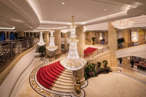 Um amplo saguão de hotel com uma escadaria de mármore, tapete vermelho e várias luminárias.