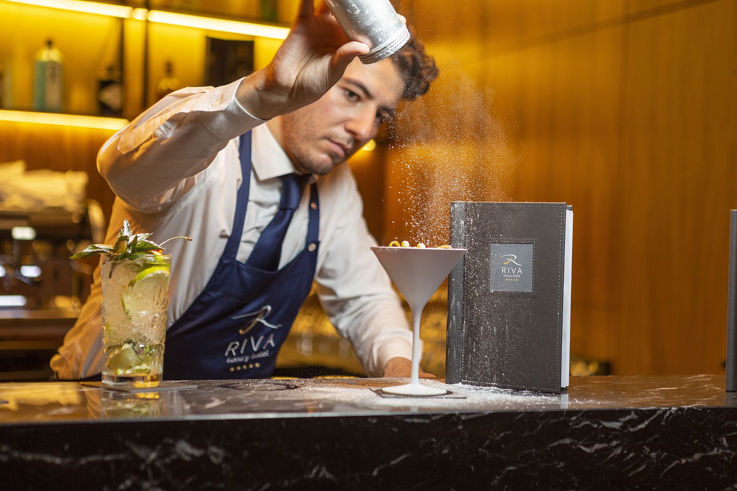 Um bartender preparando uma bebida com um coqueteleira e um copo de cocktail sobre um balcão de mármore.