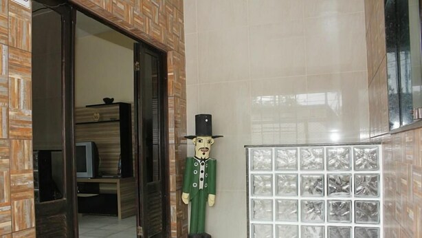 Um quarto com porta de madeira, divisória de vidro e estátua de uma figura em casaco verde e chapéu preto.
