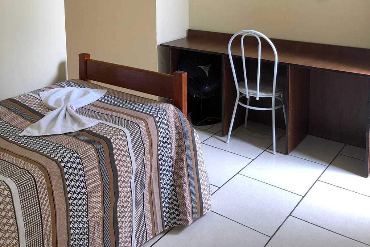 Um quarto pequeno e modesto com uma cama, uma escrivaninha, uma cadeira e uma televisão na parede.