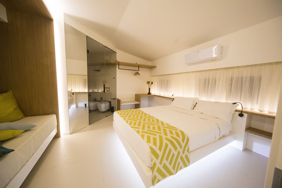 Quarto de hotel moderno com uma cama grande, um sofá, casa de banho com divisória de vidro e ar condicionado fixado na parede.