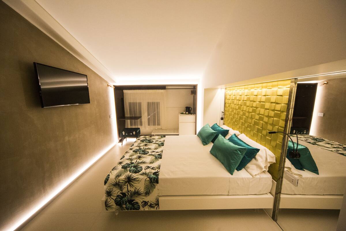 Quarto de hotel moderno com uma cama grande, televisão de tela plana na parede e banheiro visível através de uma porta aberta.