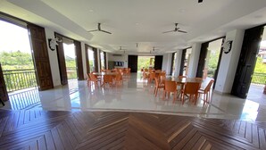 Um espaço interno amplo com piso de madeira em padrão herringbone, diversas mesas e cadeiras laranja, ventiladores de teto e grandes janelas com vista para o verde lá fora.
