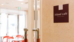 Placa com o texto 'island café', porta de madeira e cadeiras vermelhas ao fundo.
