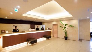 Uma área de recepção de hotel com design moderno, incluindo um balcão de atendimento, espaço de estar e uma grande planta em vaso.