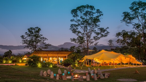 Uma pousada com um edifício bem iluminado e uma grande área coberta por tendas, cercada por árvores e um cenário montanhoso.