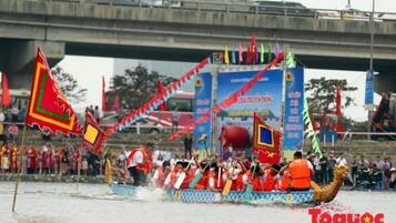 Uma corrida de barcos dragão com participantes em um barco, espectadores observando da margem e uma ponte ao fundo.