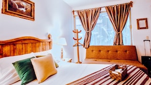 Quarto de hotel com cabeceira de madeira, uma cama grande com almofadas brancas e verdes, tapete listrado, porta-roupas de madeira e uma janela com cortinas.