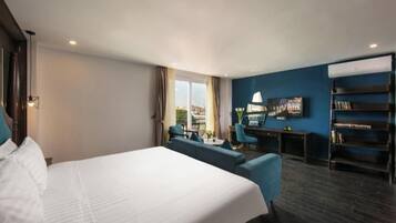 Quarto de hotel com uma cama grande, área de estar com escrivaninha e uma estante de livros.