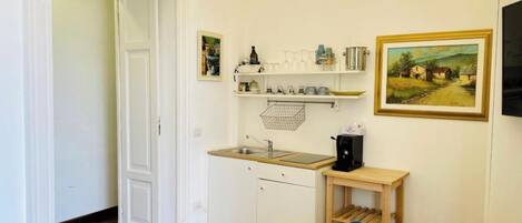 Uma cozinha compacta com armários brancos, bancada de madeira e uma pia. Há uma pequena mesa com uma cafeteira e uma cesta na parede.