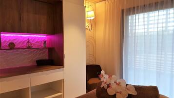 Um espaço de spa com uma mesa de massagem, um armário de madeira com uma prateleira iluminada em rosa, uma luminária de parede e uma janela com cortinas leves.