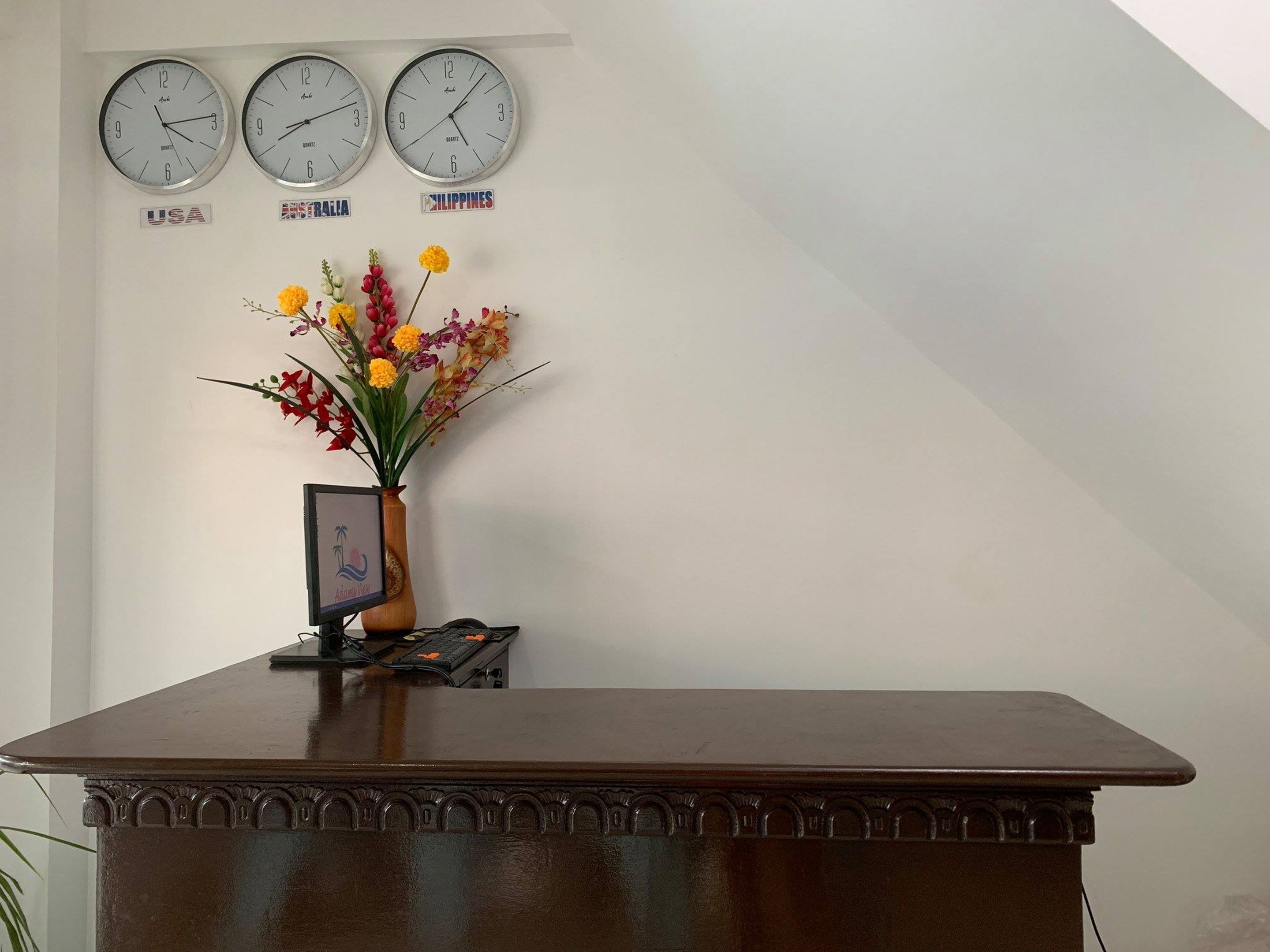 Um balcão de atendimento com tampo de madeira, um vaso com flores e três relógios na parede com as indicações EUA, Austrália e Filipinas.