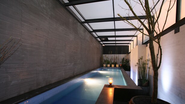 Piscina interna com piso ladrilhado, teto de vidro e uma pequena mesa com abajur.