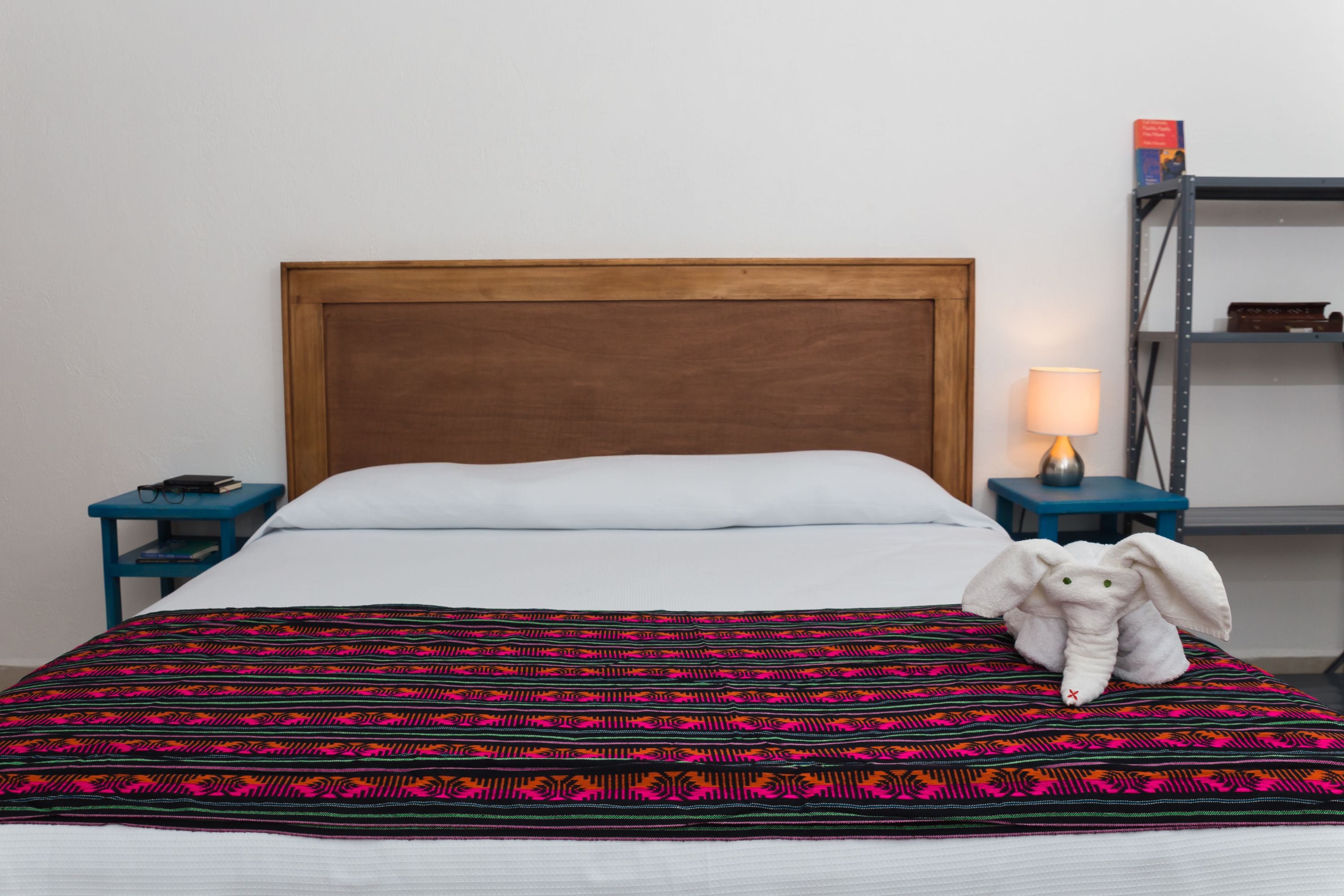Uma cama bem arrumada com um cobre-leito estampado, cabeceira de madeira, abajur e uma pequena prateleira com um livro e uma luminária.