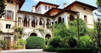 Um edifício de dois andares com janelas em arco, um jardim com diversas plantas e um caminho de cascalho.