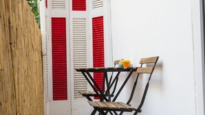 Um pátio exterior estreito com mesa e cadeiras de madeira, vedação de bambu e paredes brancas com persianas vermelhas.