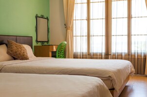 Quarto com duas camas, uma cadeira verde, uma mesa de cabeceira de madeira, um espelho e uma janela com cortinas.