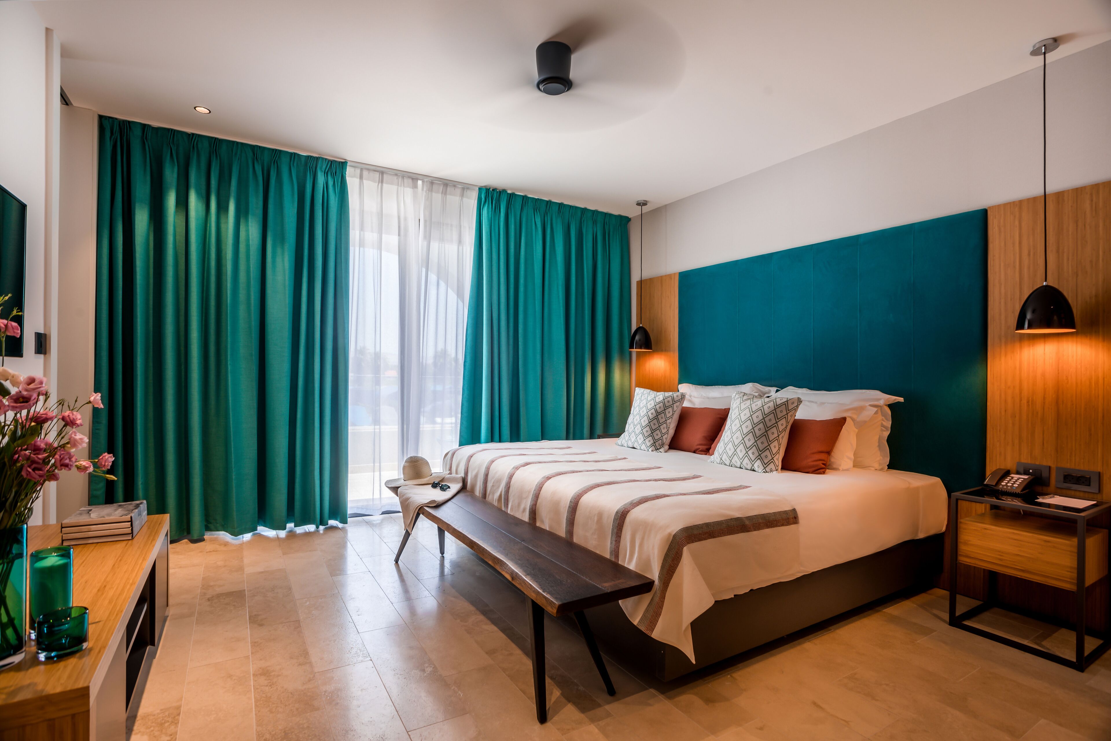 Quarto de hotel moderno com uma cama grande, um banquinho, uma mesinha de cabeceira com abajur e vista da cidade por grandes janelas.