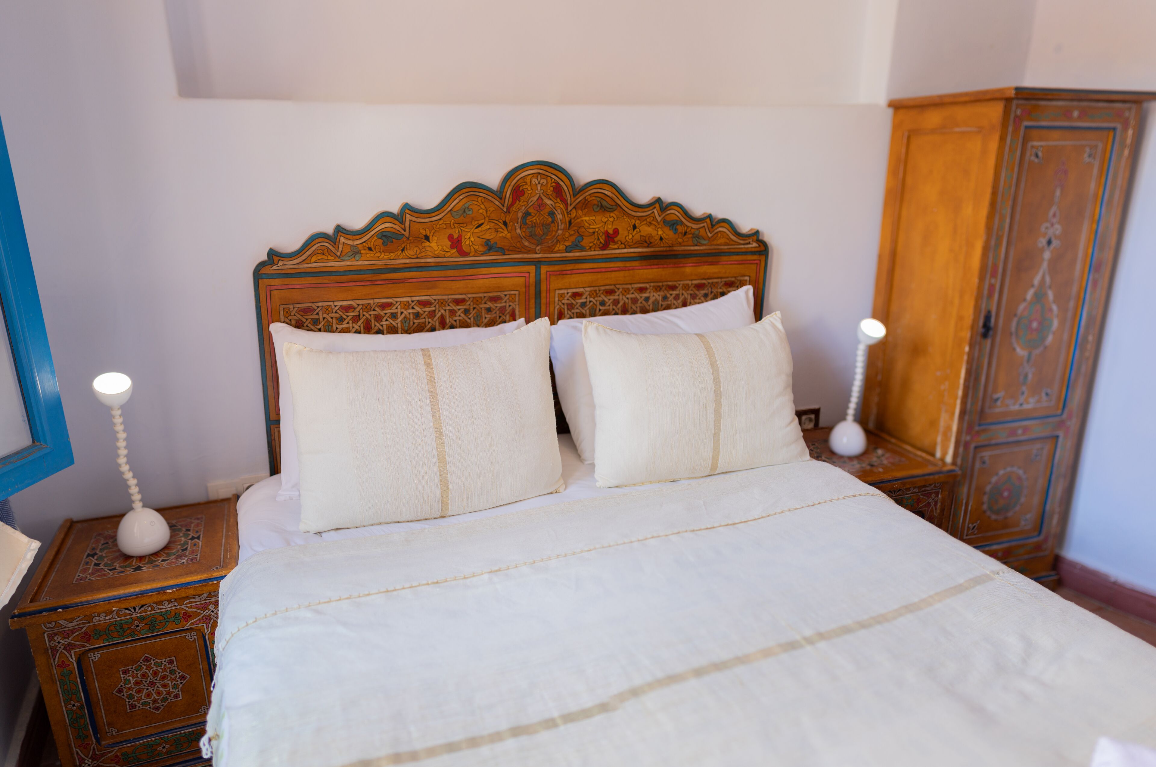 Uma cama com cabeceira decorativa, duas mesas de cabeceira com abajures e um guarda-roupa de madeira.