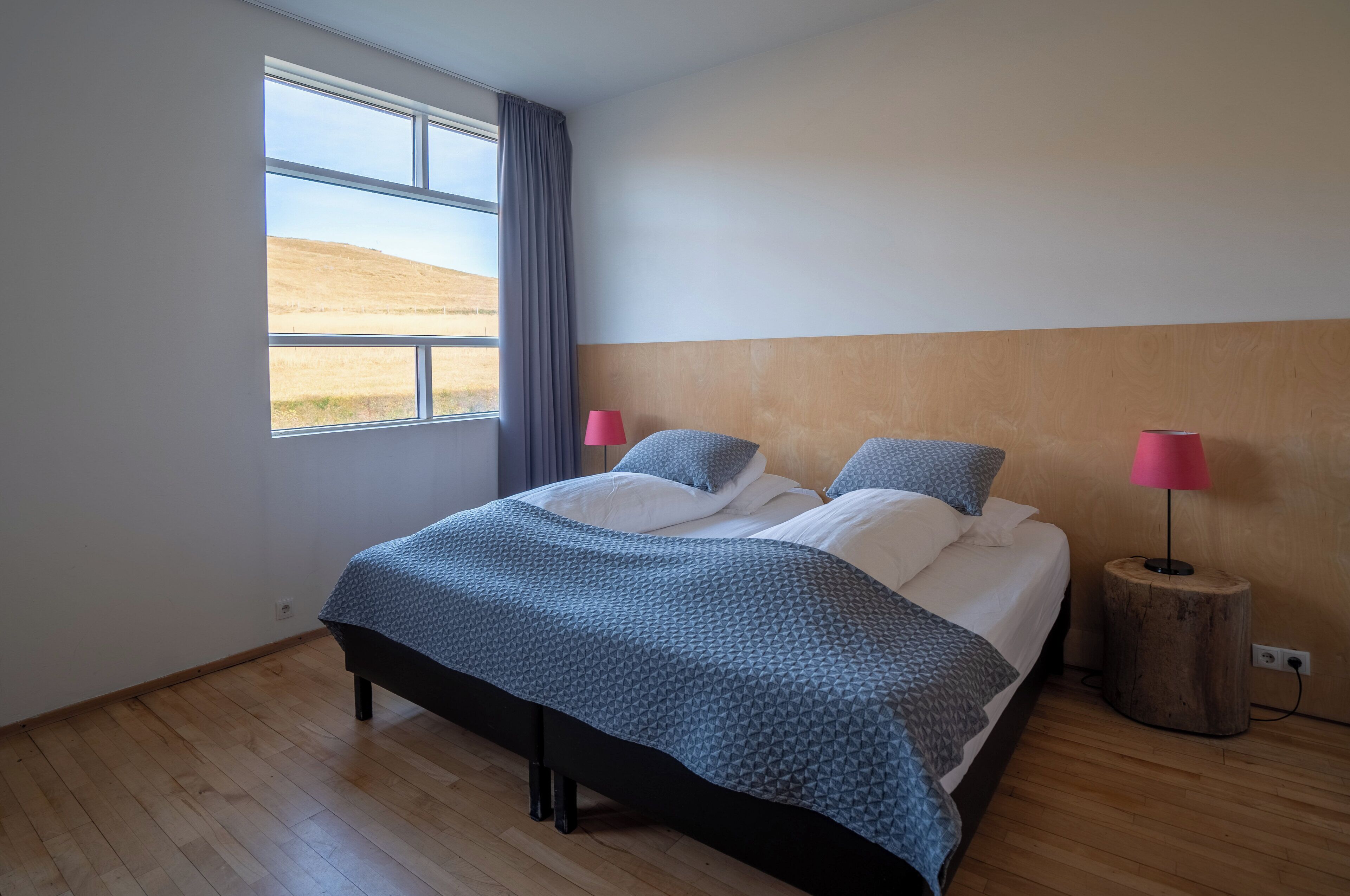 Quarto com cama, duas luminárias de cabeceira, janela com vista para uma colina e cabeceira de madeira.