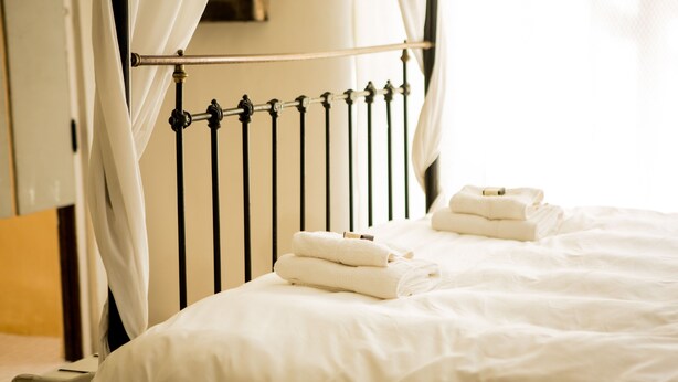 Cama bem arrumada com lençóis e toalhas brancas, cabeceira de metal e mesa de cabeceira com abajur.