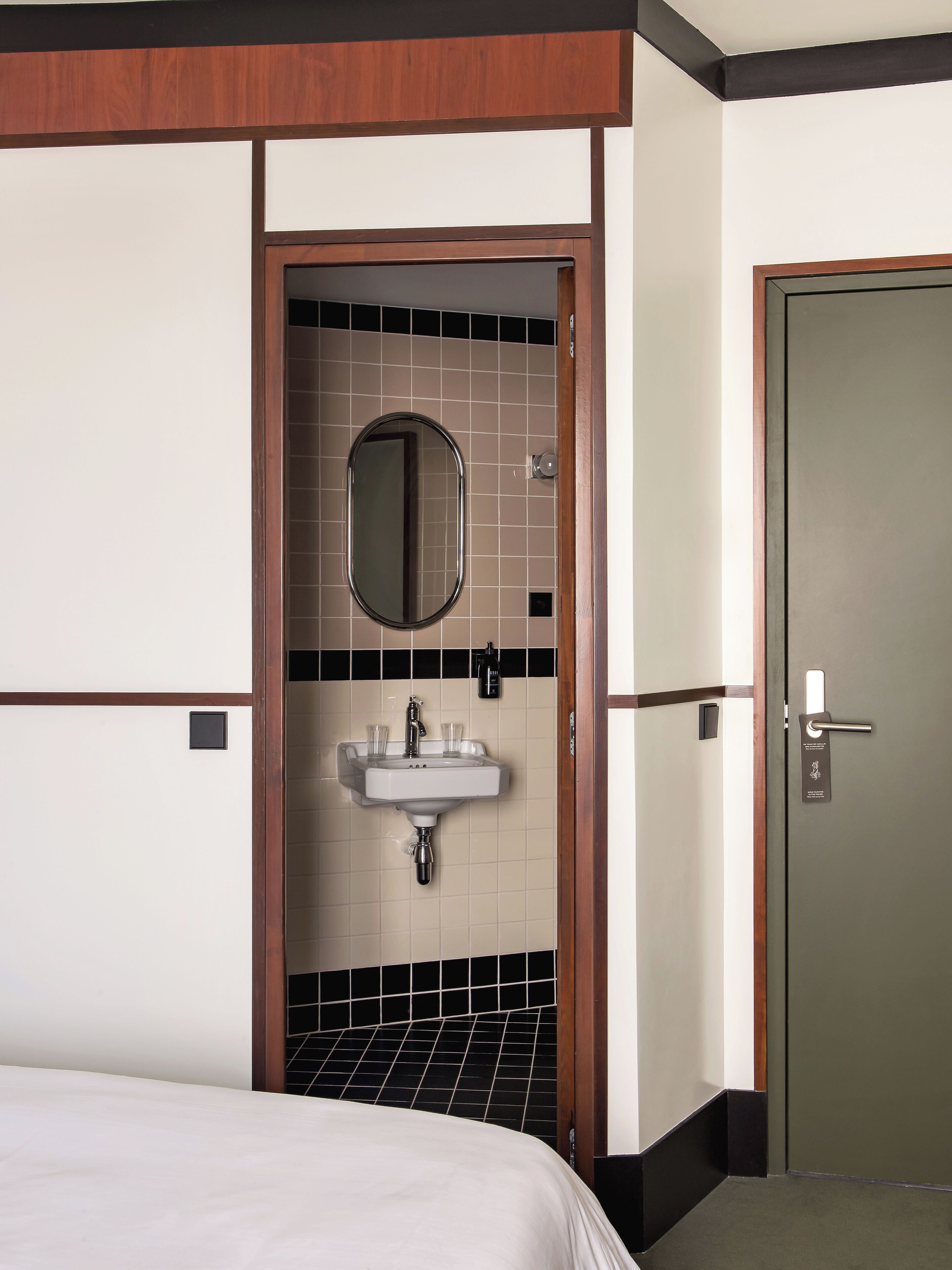 Quarto de hotel com uma cama, pia com três copos, espelho e uma porta.
