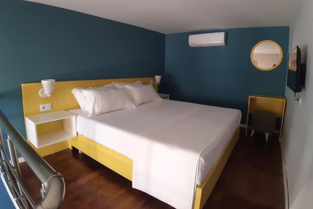 Quarto de hotel com cama, mesas de cabeceira, espelho e aparelho de ar condicionado.