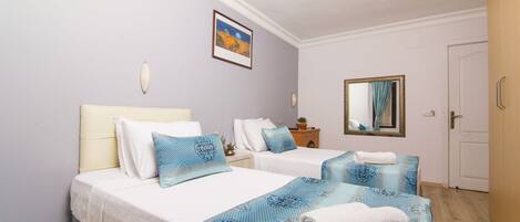 Quarto de hotel com duas camas, cada uma com colcha estampada azul e travesseiros brancos. Há uma pequena mesa com uma planta, um espelho e uma cortina azul, e uma porta à direita.