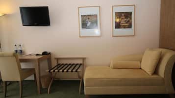 Quarto de hotel com sofá, escrivaninha com cadeira, televisão fixada na parede e duas imagens emolduradas.