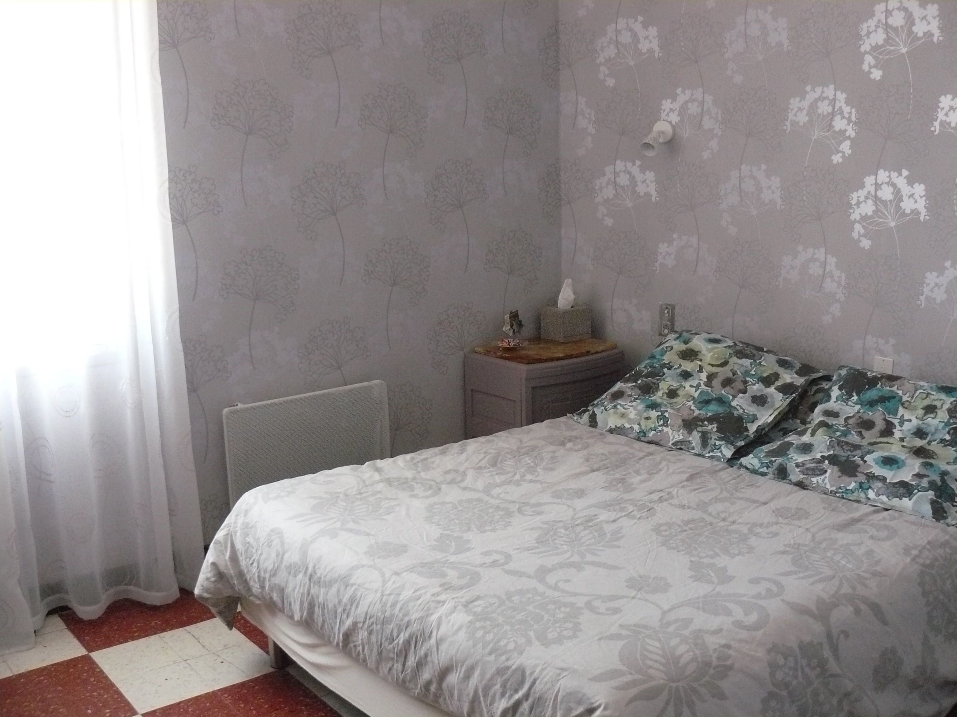 Quarto com cama, mesa de cabeceira, radiador e janela com cortinas leves.