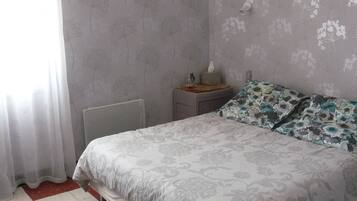 Quarto com cama, mesa de cabeceira, radiador e janela com cortinas leves.