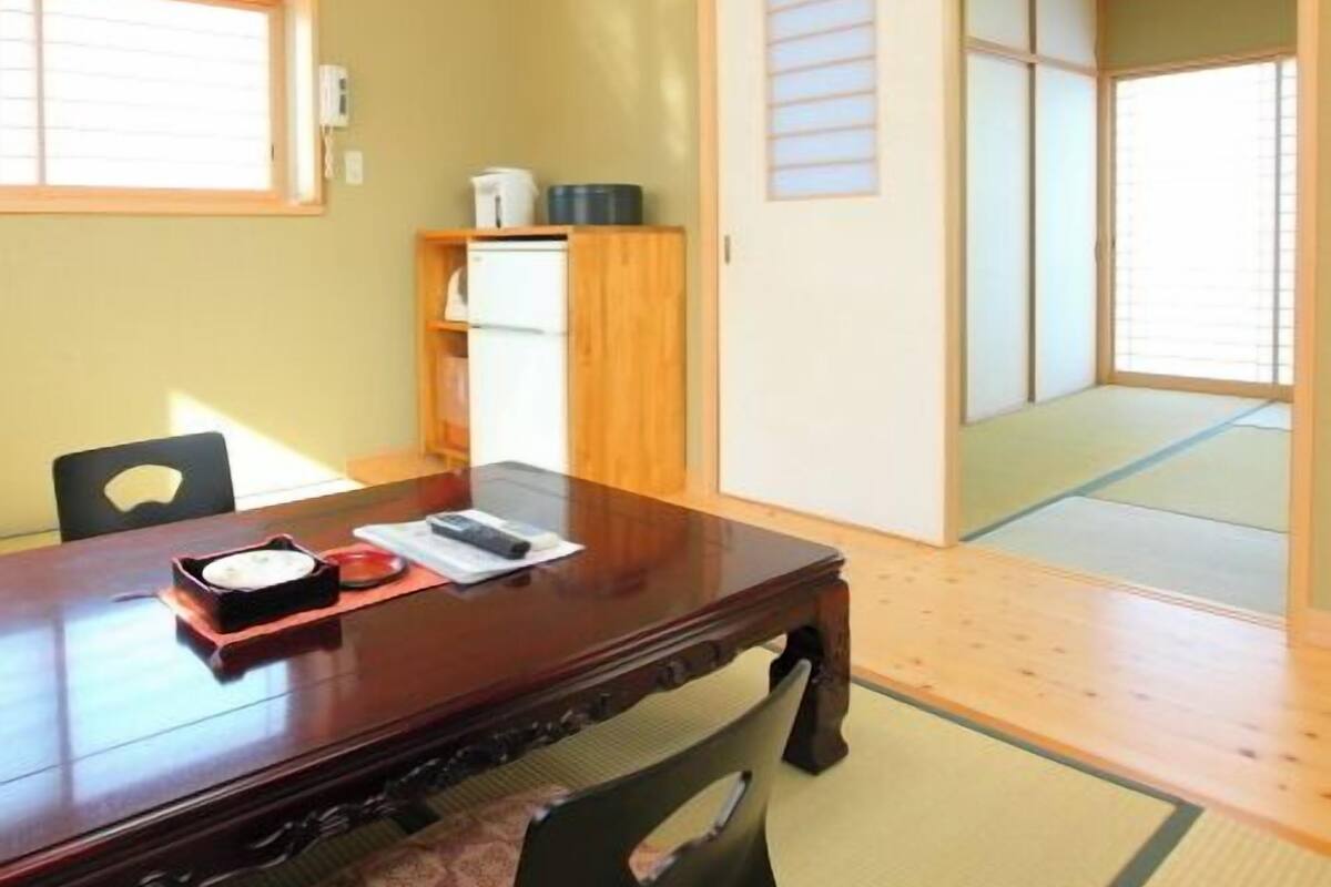 Quarto japonês tradicional com tatames, uma mesa de madeira, um frigorífico e uma porta deslizante.