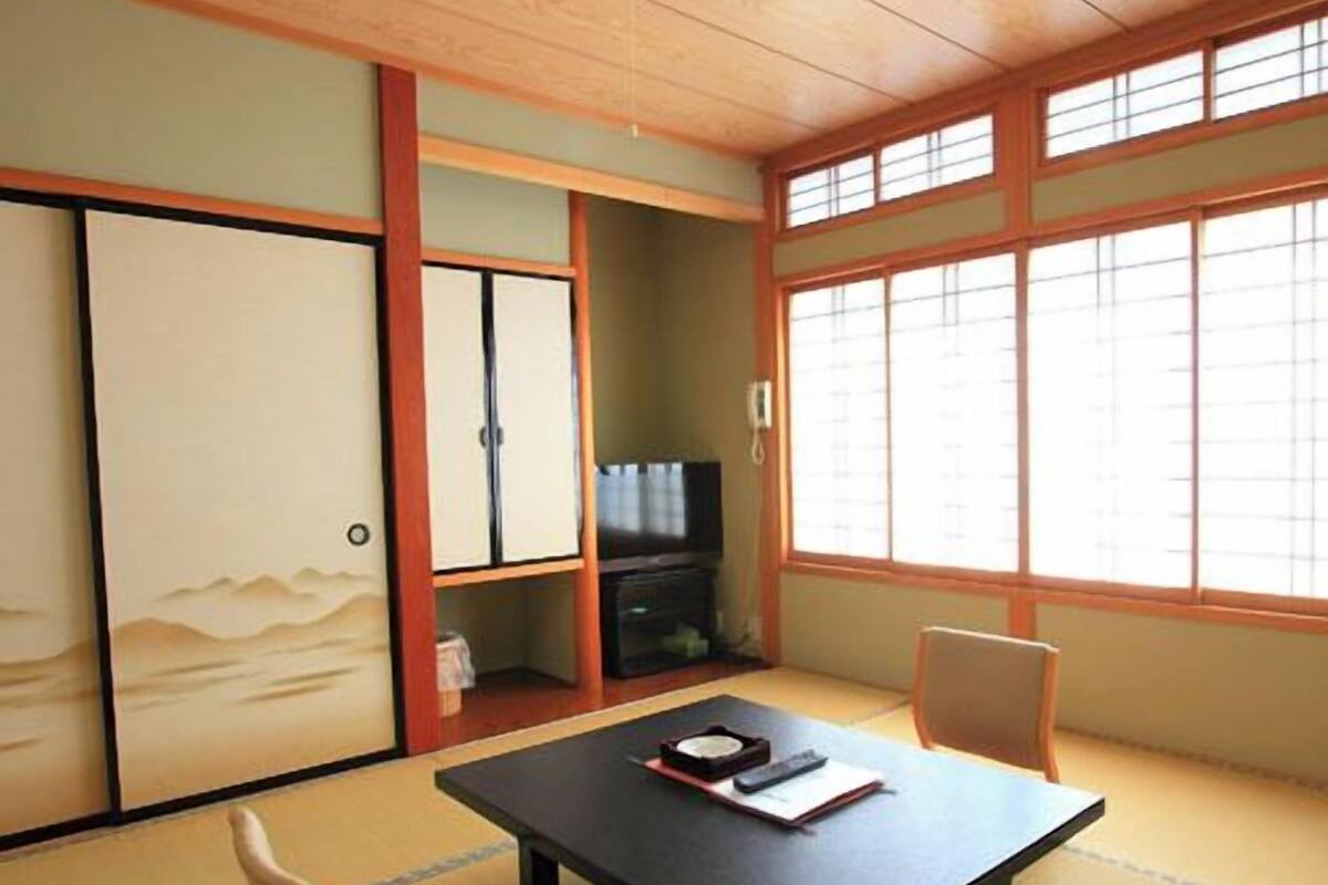 Um quarto japonês tradicional com tatames, uma mesa baixa com almofada, uma televisão e portas deslizantes.