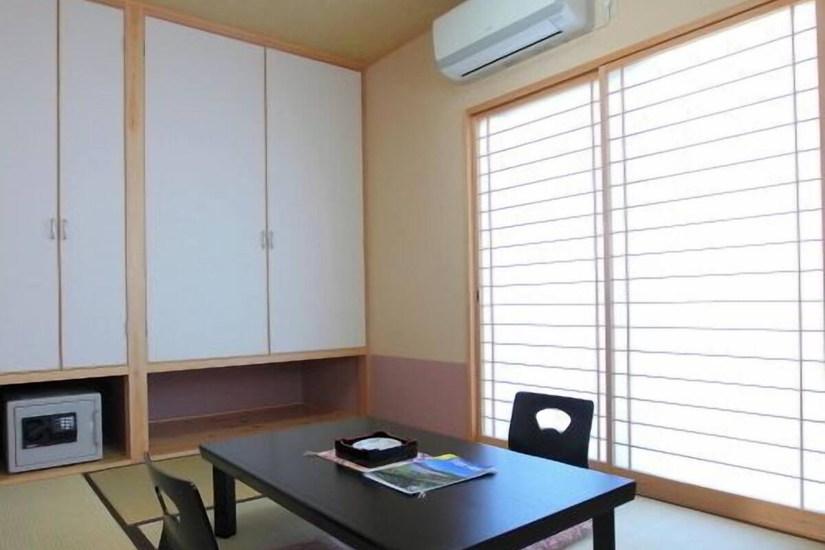 Quarto com piso de tatame, uma mesa preta com cadeiras, janela com persianas e um ar condicionado.