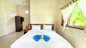 Cama bem arrumada com lençol branco e um enfeite de cisne de toalha azul, mesa de cabeceira de madeira com uma flor e uma janela com cortinas brancas.