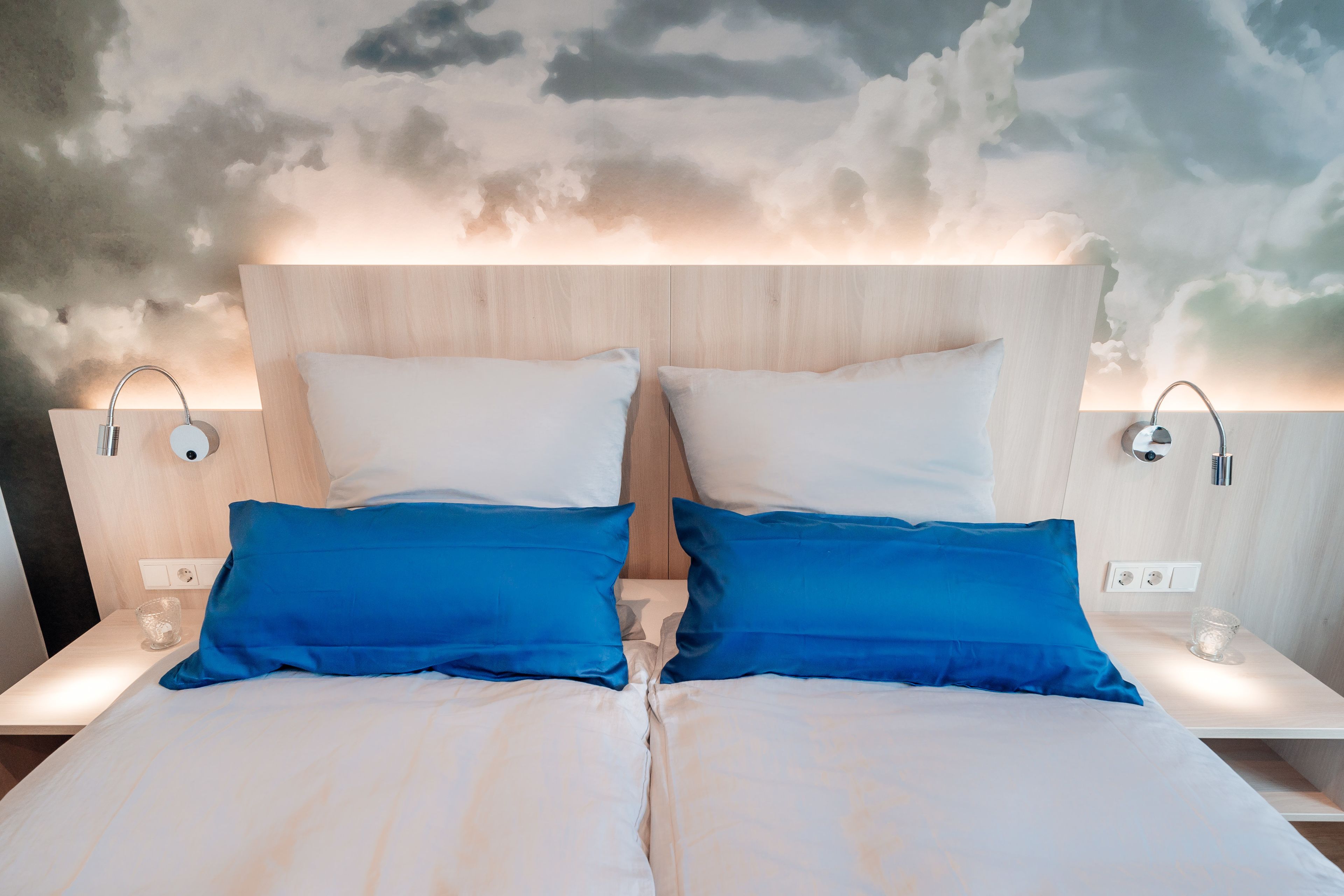 Cama com roupa de cama branca e azul, cabeceira de madeira e parede com mural de nuvens.