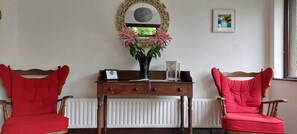 Quarto com duas cadeiras vermelhas estofadas, uma mesa consola de madeira com um vaso de flores cor-de-rosa, um quadro emoldurado e um espelho com moldura decorativa.