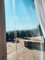Vista da janela com céu claro, área de varanda e colinas e árvores ao longe.