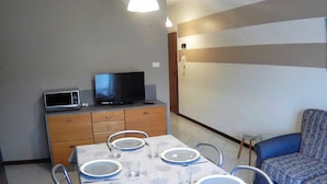 Uma pequena sala de jantar com uma mesa posta para quatro pessoas, micro-ondas, televisão em um armário e uma poltrona estampada.