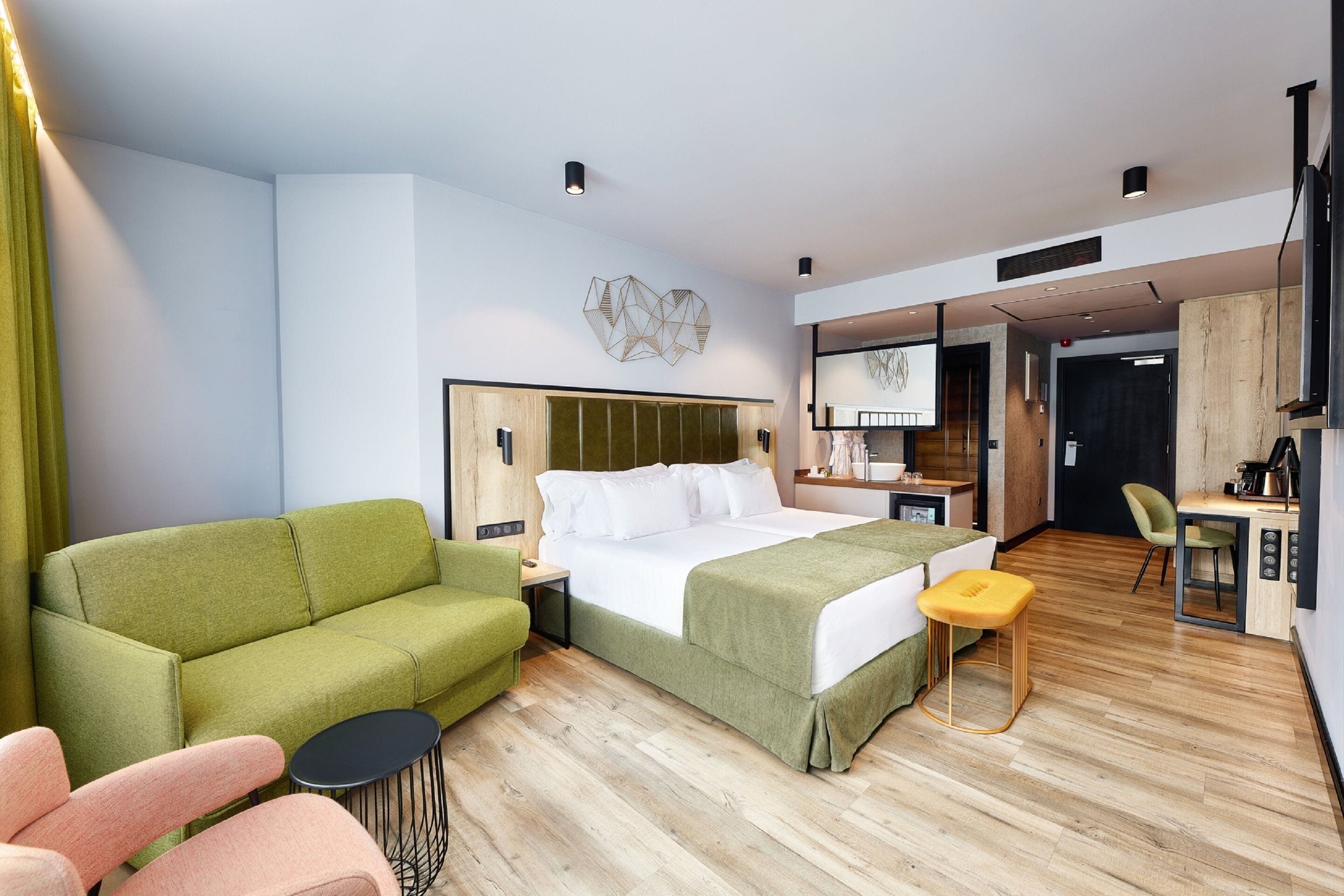 Quarto de hotel moderno com um sofá verde, uma cama com roupa de cama branca, uma mesa redonda pequena e um banquinho amarelo.