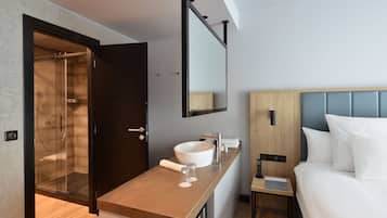 Quarto de hotel moderno com toucador de madeira, um espelho grande, uma cama com roupa de cama branca e uma pequena mesa de cabeceira.