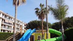 Um hotel com um escorregador azul e verde no pátio, cercado por palmeiras e um edifício amarelo.