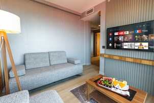 Sala de estar moderna com sofá cinzento, mesa de centro de madeira com frutas e bebidas, e uma TV fixada na parede exibindo diversos serviços de streaming.