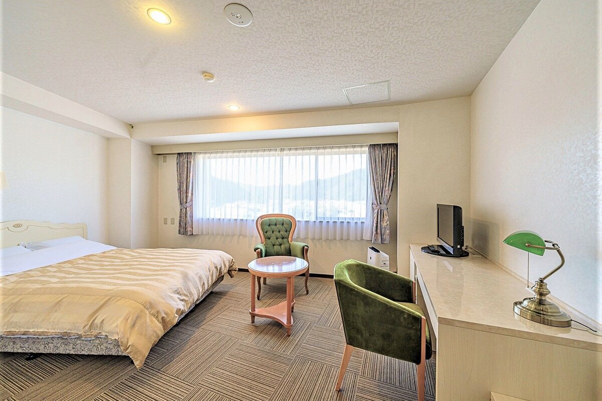 Quarto de hotel com uma cama, uma escrivaninha com computador, uma cadeira e uma televisão.