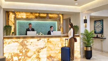 Balcão de recepção de hotel com tampos de mármore, duas recepcionistas, um hóspede com mala e decoração moderna, incluindo um candelabro e obras de arte.