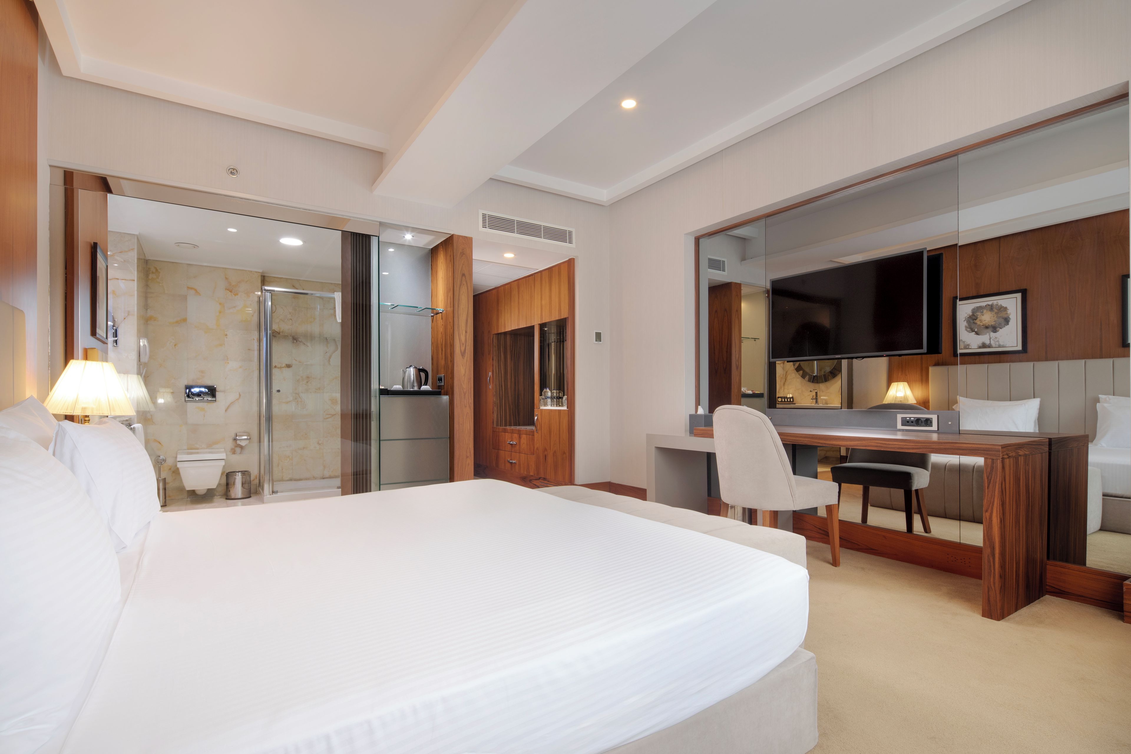 Quarto de hotel com cama grande, escrivaninha, cadeira, televisão e banheiro com chuveiro e vaso sanitário.