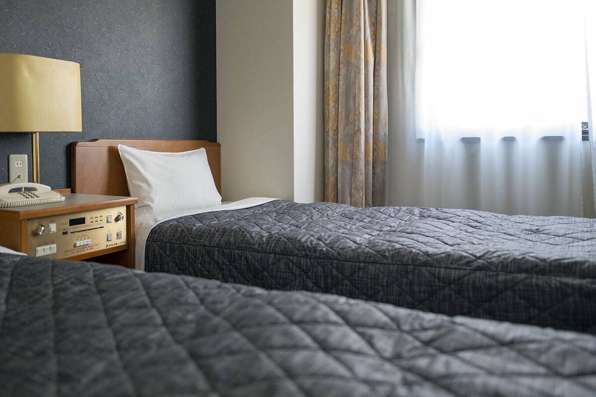 Quarto de hotel com duas camas, uma mesa de cabeceira com abajur, telefone e janela com cortinas.