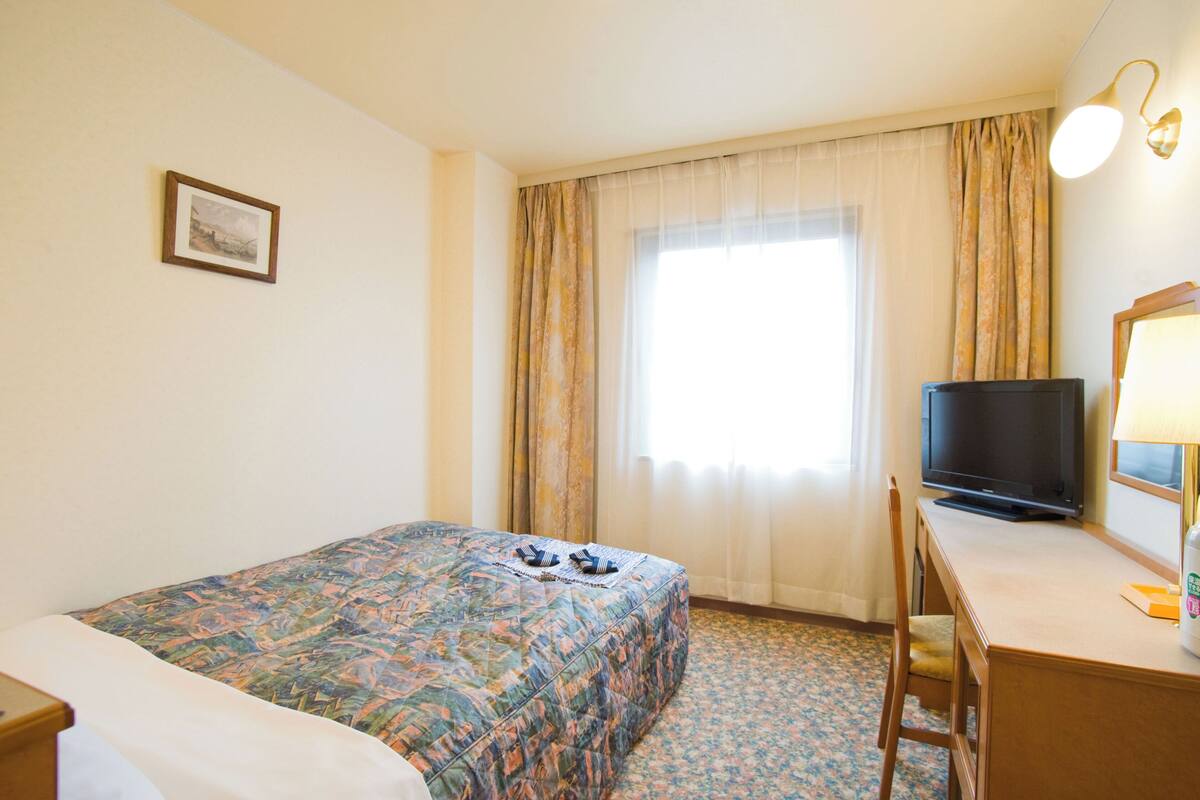 Quarto de hotel com cama, escrivaninha, televisão e janela com cortinas.