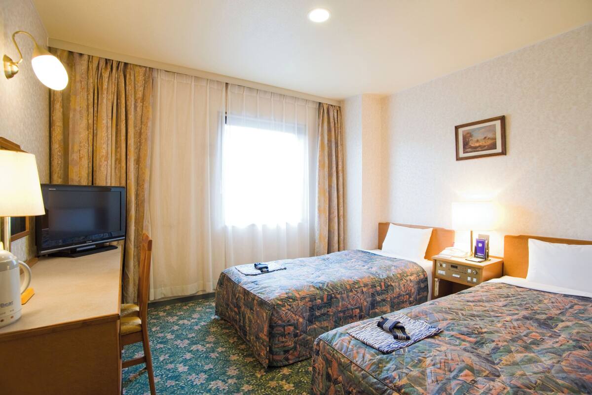 Quarto de hotel com duas camas, televisão, secretária com candeeiro e janela com cortinas.